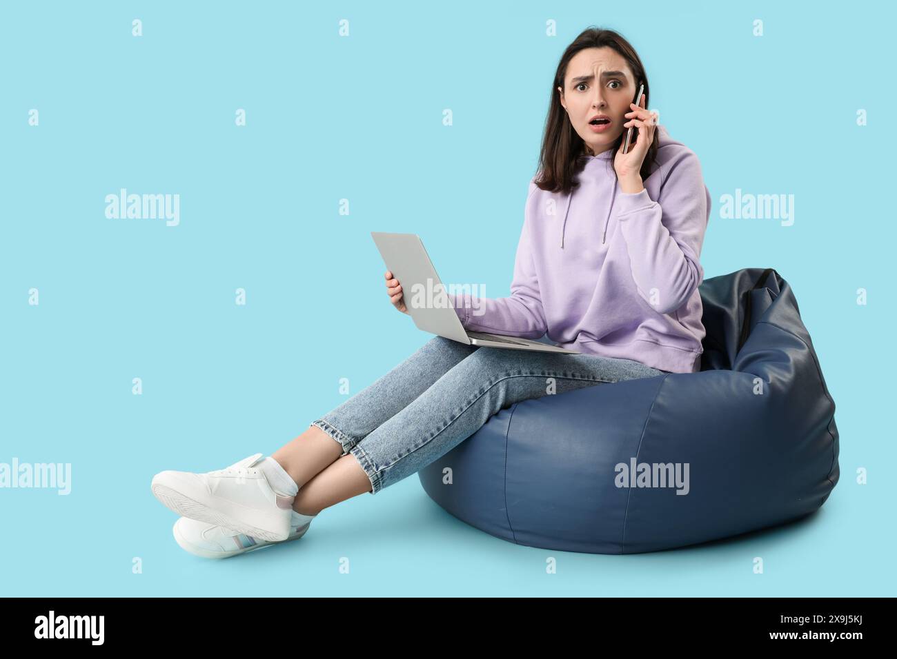 Shocked young woman with laptop talking by phone while sitting on bean ...