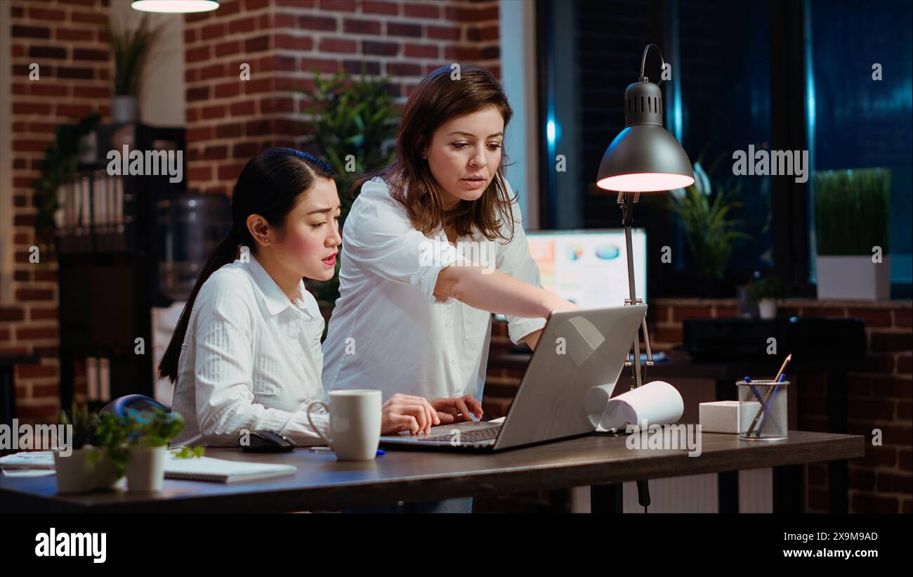 Asian staff member calling manager to her computer desk to ask for feedback on project progress ...