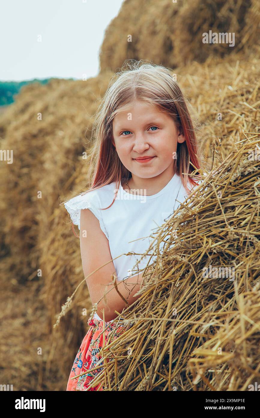 Young Girl With Blonde Hair In A White Top Stands Near A Haystack In The Countryside On A Summer
