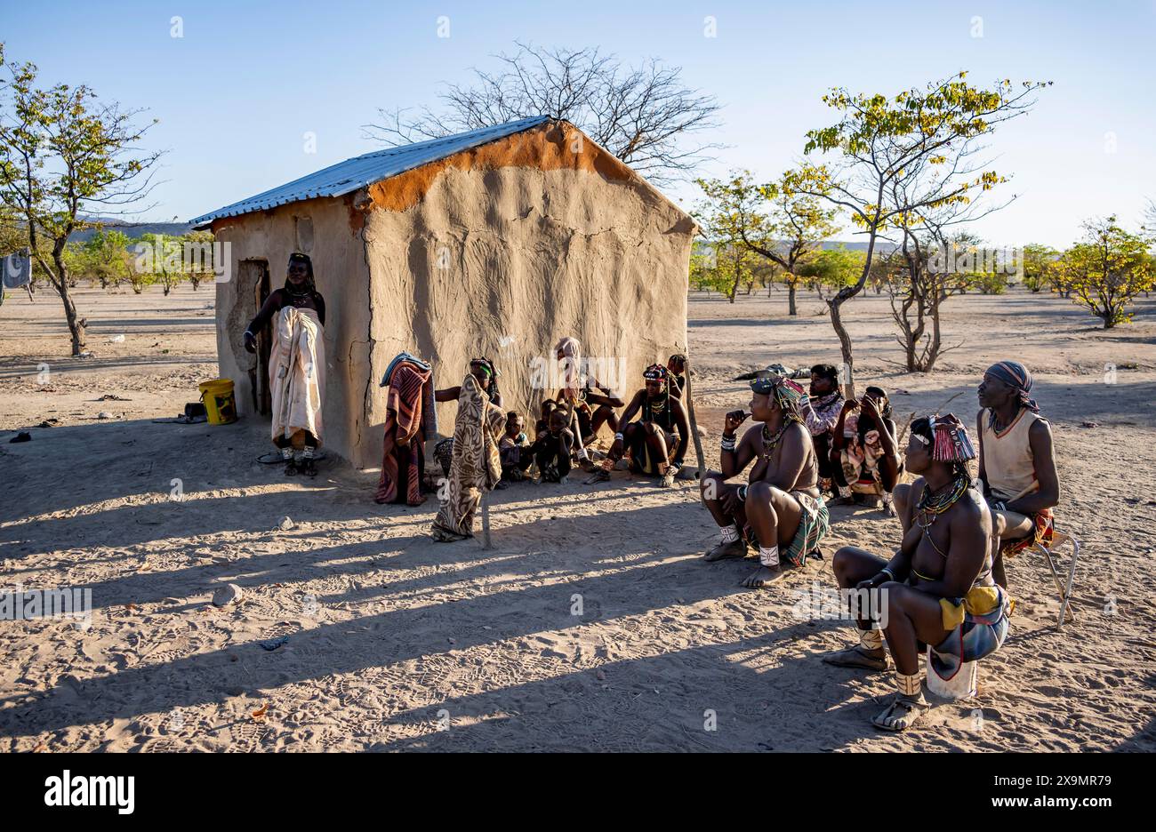 Traditional Hakaone woman and men sit in front of a mud house in their ...