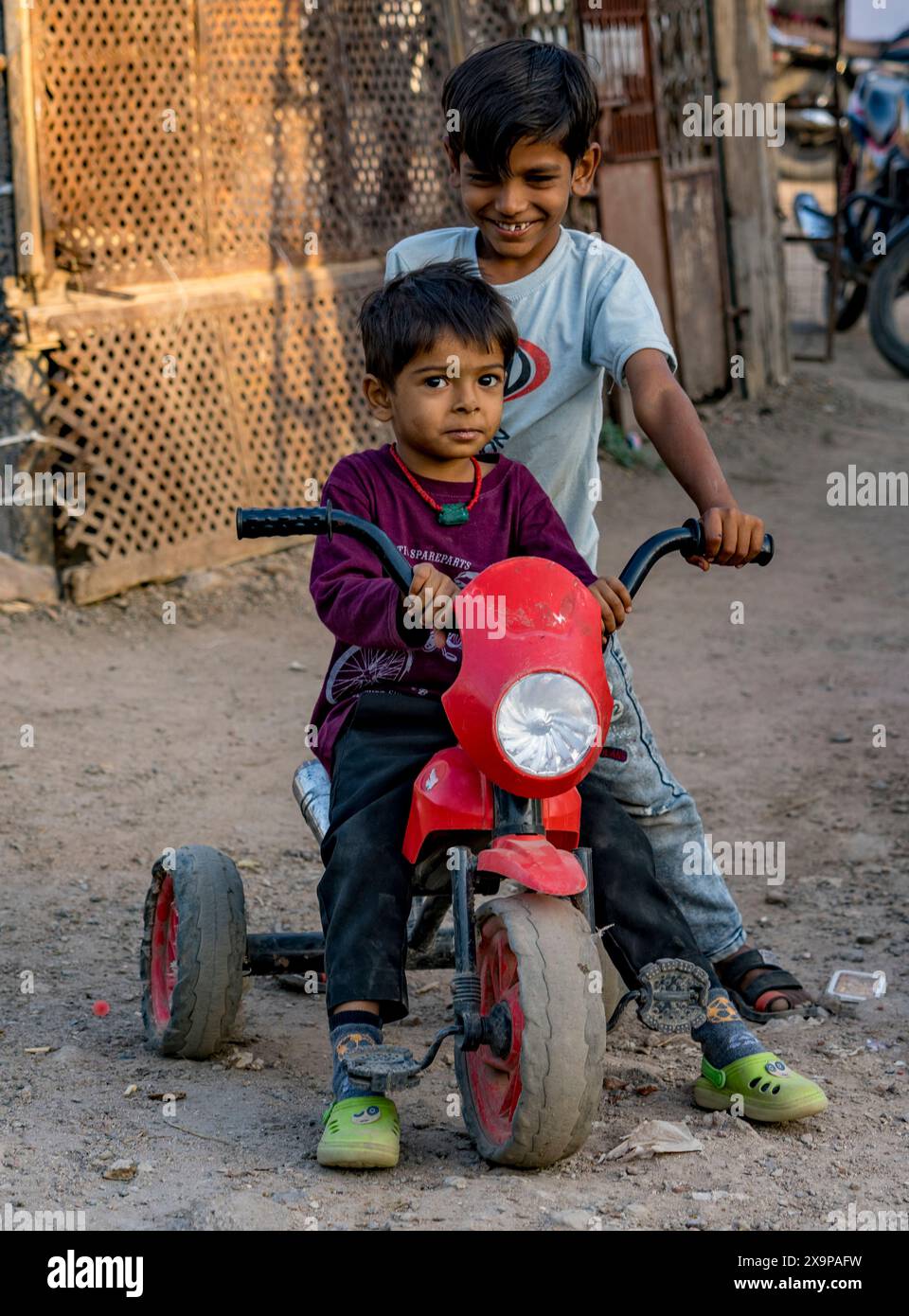 Older brother helps younger sibling ride a red tricycle on a dirt road ...