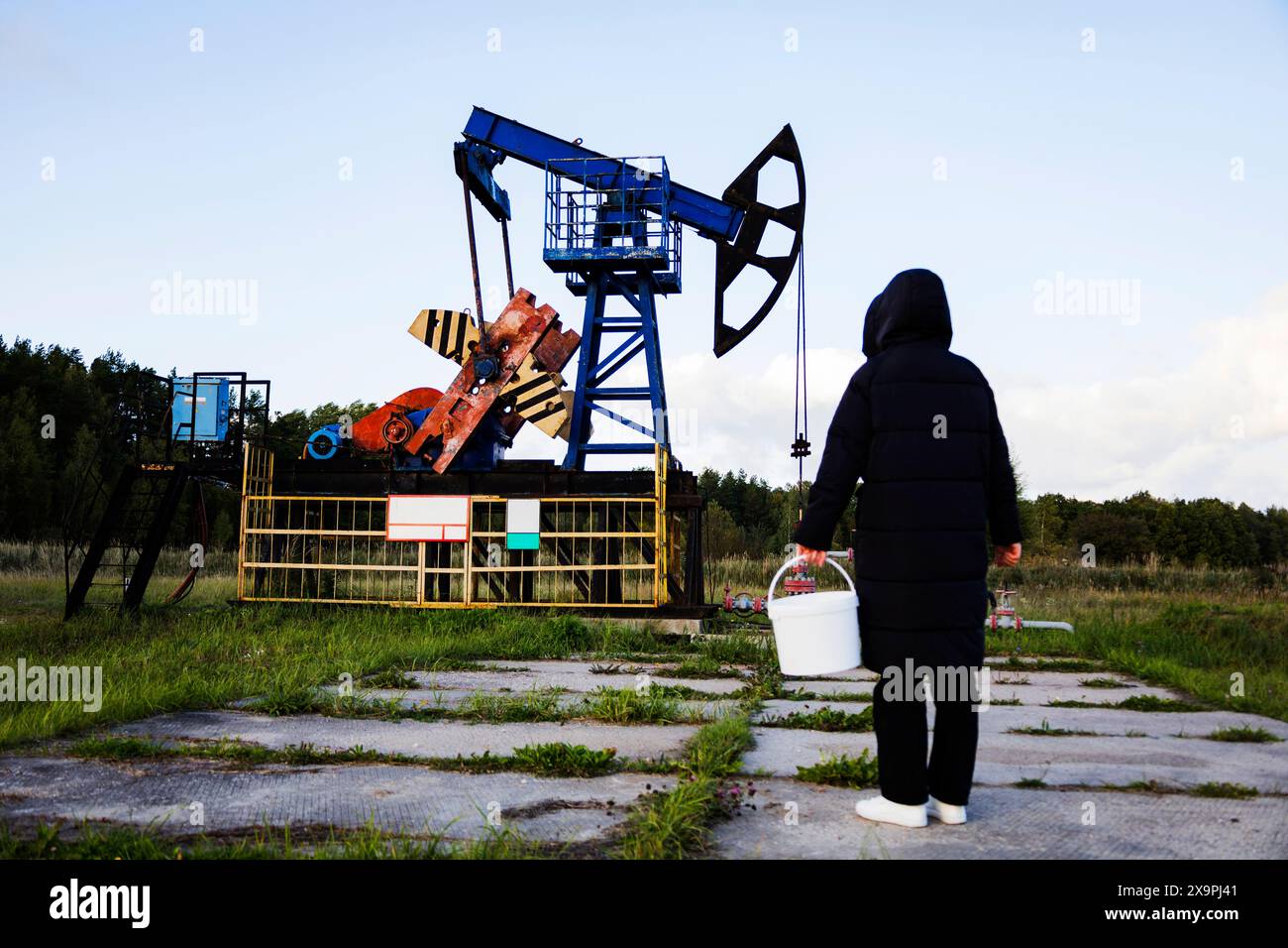 A woman wearing a black coat stands by an oil pump. The background ...