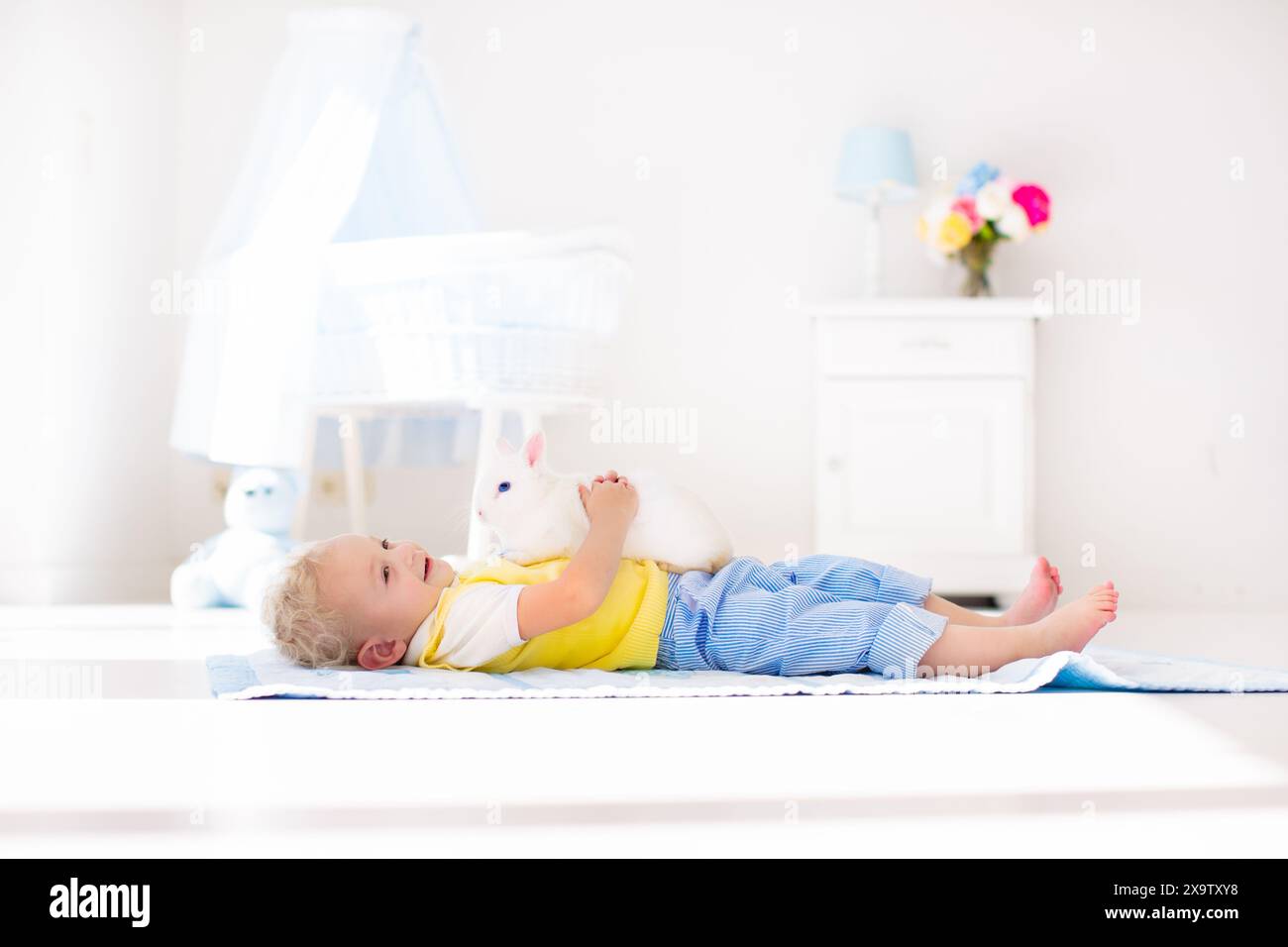 Cute blond curly baby boy playing with rabbit in a white sunny bedroom ...