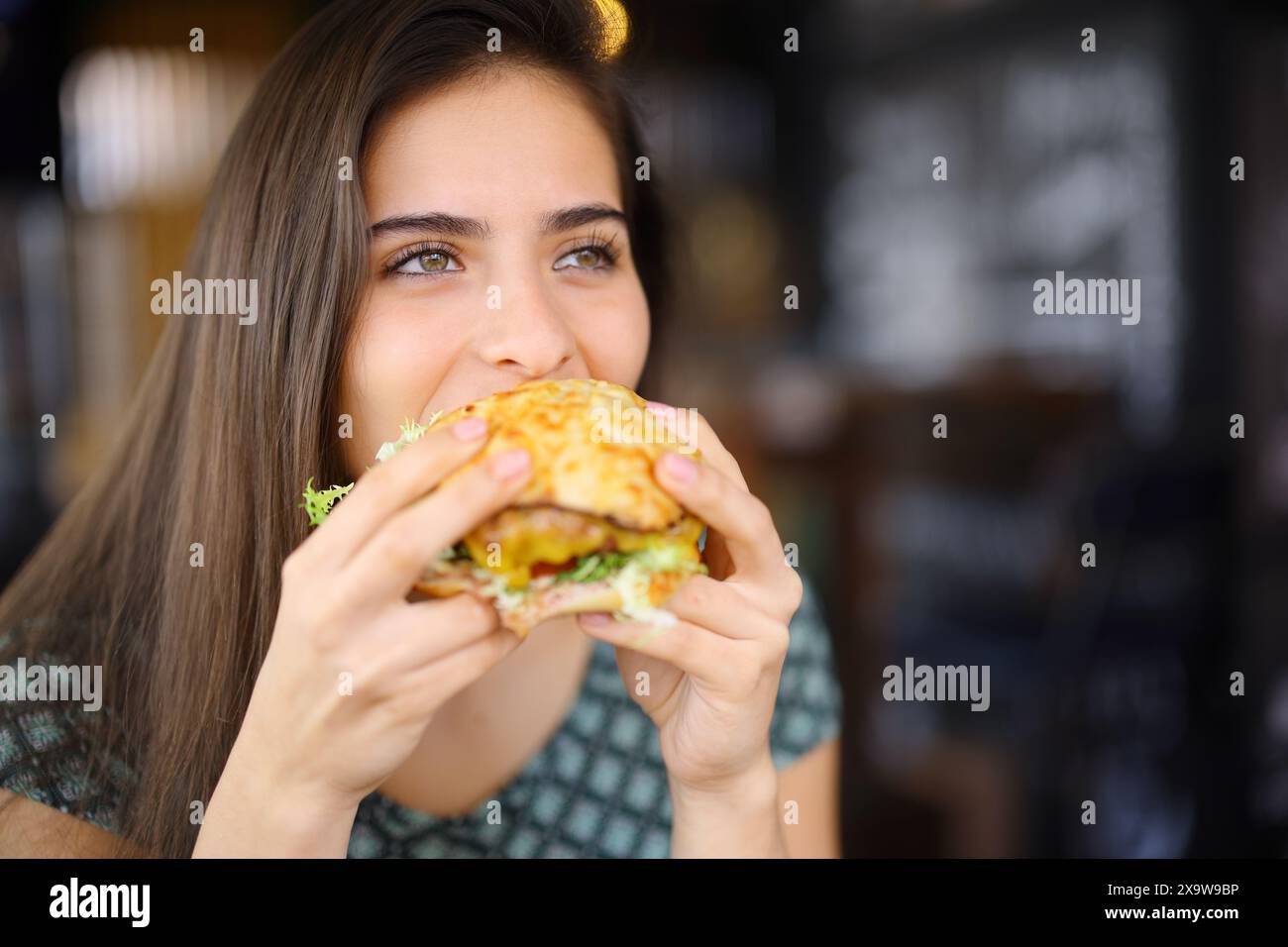 Happy woman eating hamburger in a restaurant interior looking away ...