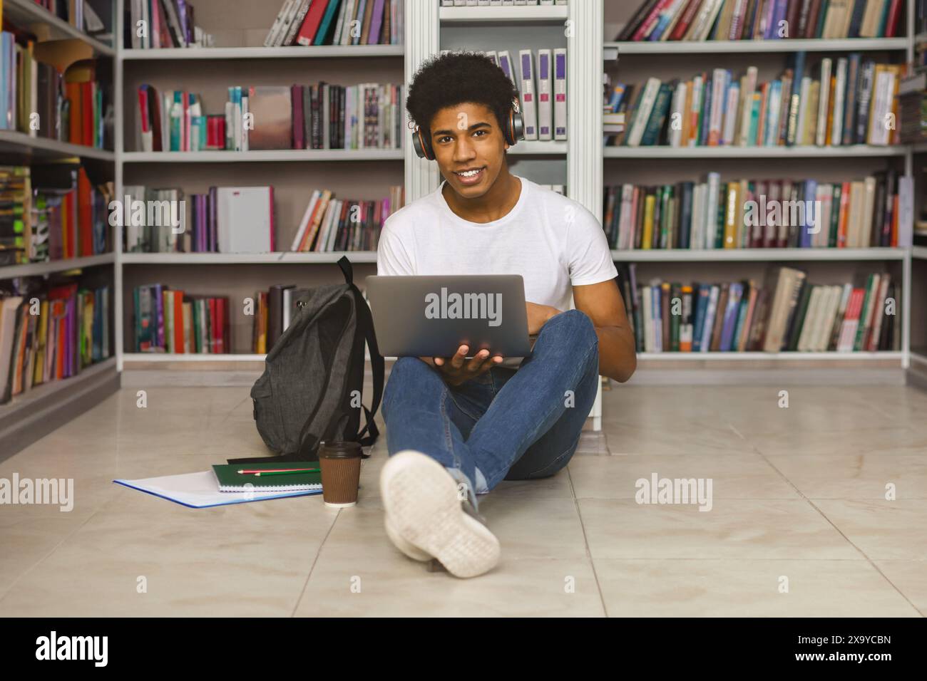 Afro student with headset leaning on bookshelf and using laptop Stock ...