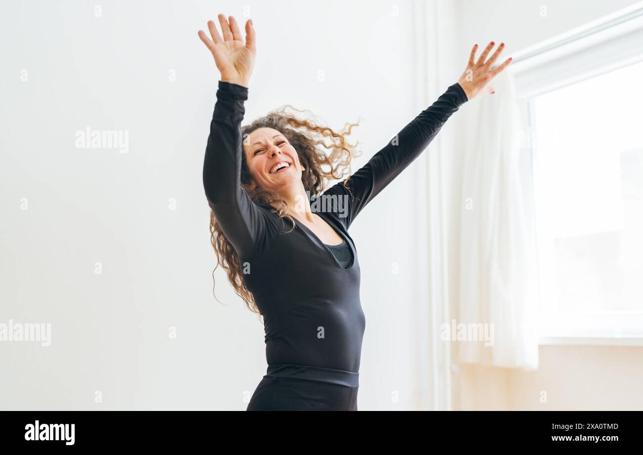Joyful woman in black outfit captured mid-dance in bright room arms ...