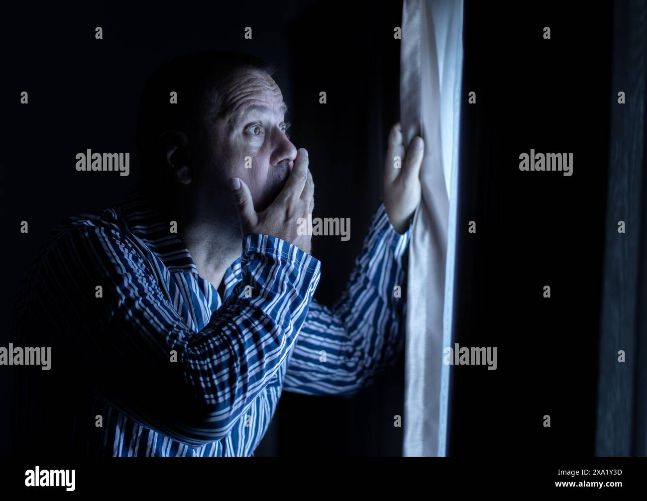 Frightened man in striped pajamas looks out behind curtained window, at ...