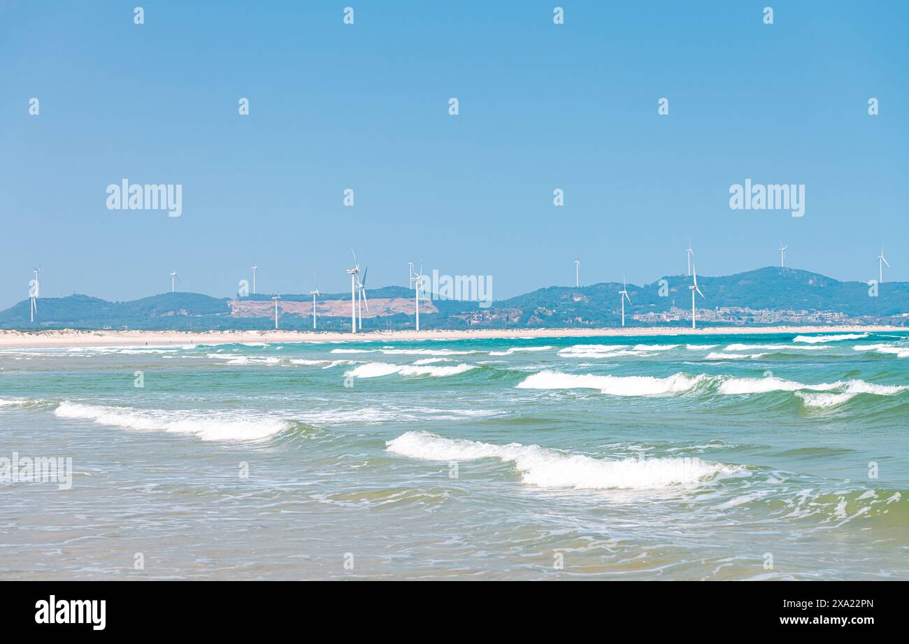 A scenic view of wind turbines line a sandy beach with waves crashing ...