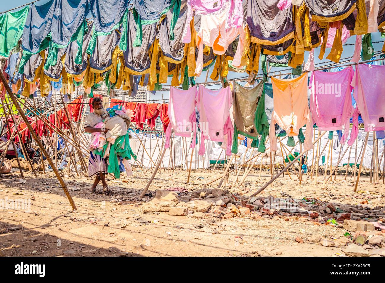 A man carrying laundry under vibrant hanging clothes in Mumbai sunlight ...