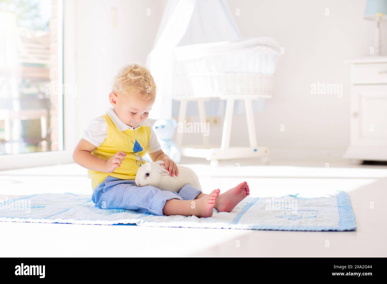 Cute blond curly baby boy playing with rabbit in a white sunny bedroom ...