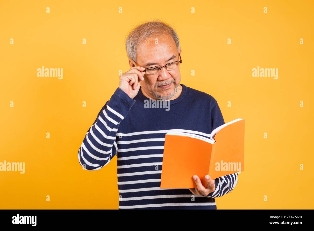 Portrait smiling Asian old man wearing glasses reading a book studio shot isolated yellow ...