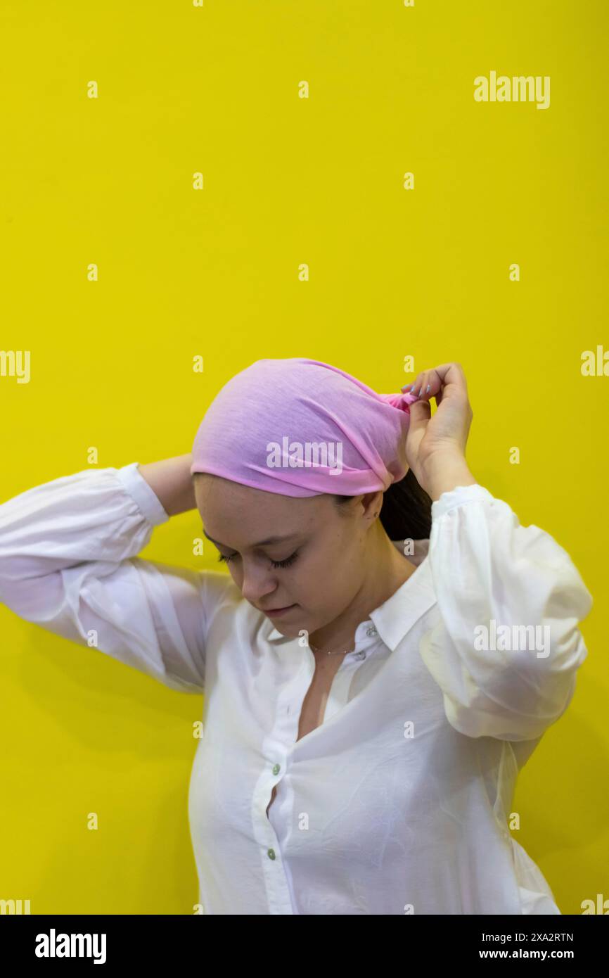 Female cancer patient putting on a pink headscarf on yellow background ...