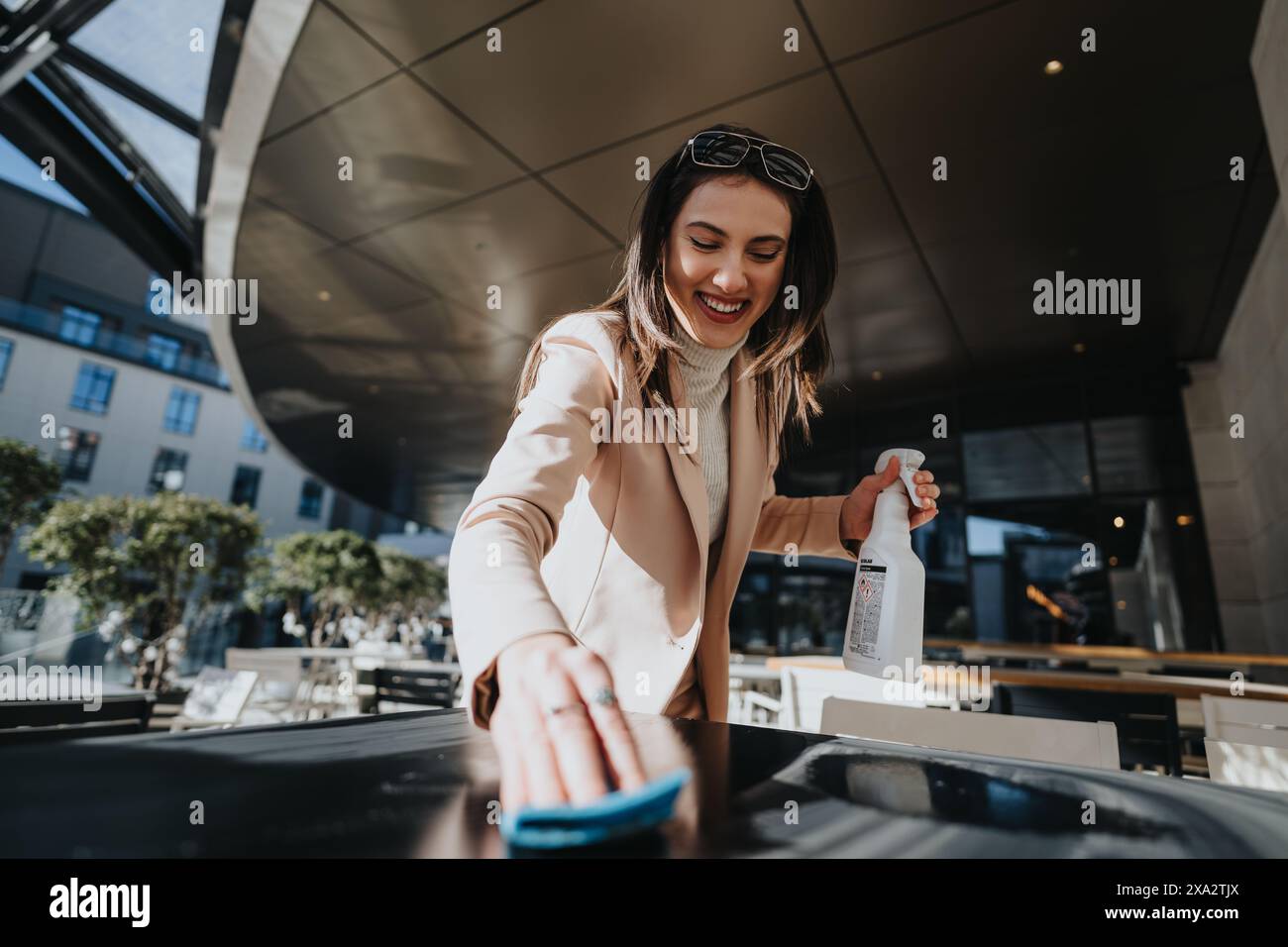 Joyful female employee sanitizing outdoor dining table with ...
