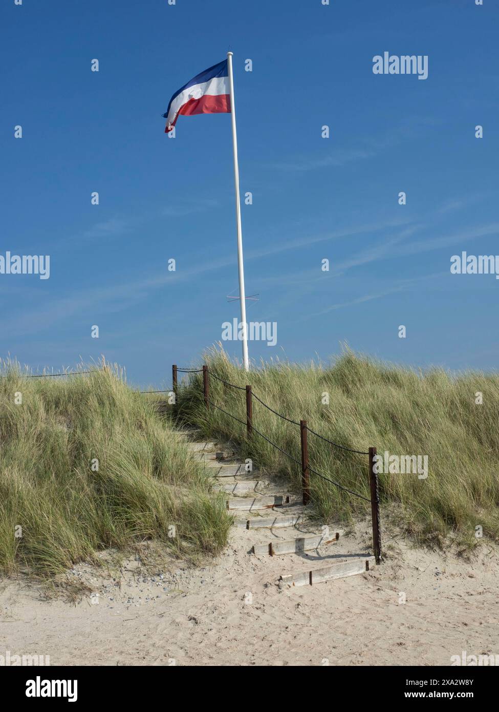 A staircase leads through grassy dunes to a high-flying flag, the sky ...