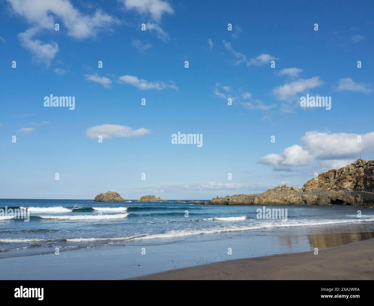 A vast beach with small waves, rocks behind it and a clear sky with few ...