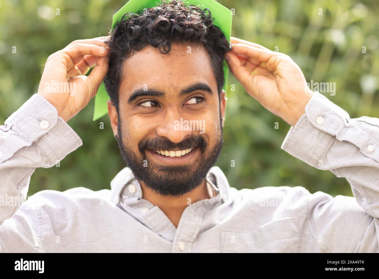 A smiling man with curly hair is putting on a green paper hat in a ...
