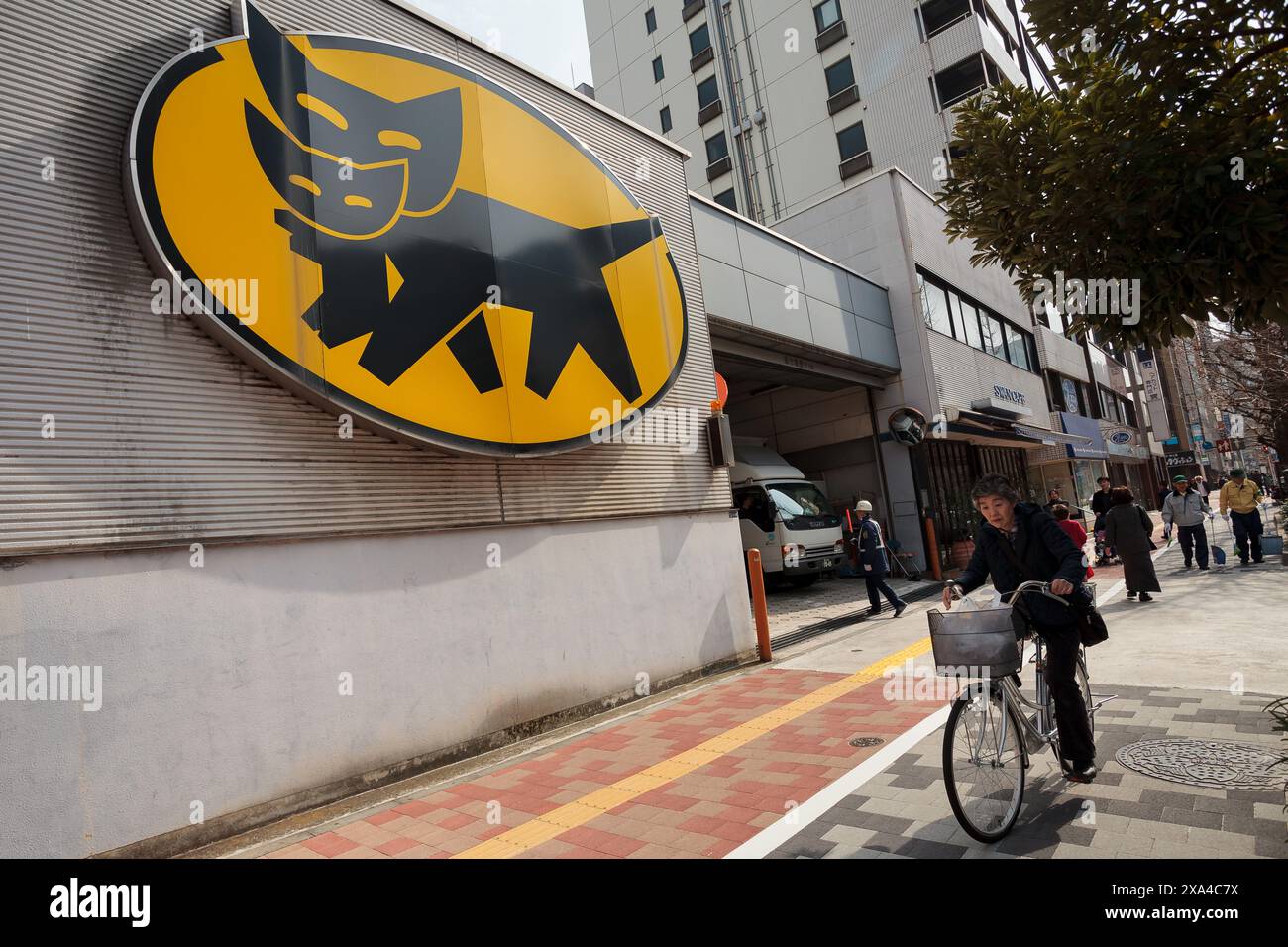 A woman rides a bicycle past a large logo for Yamato Delivery Company ...
