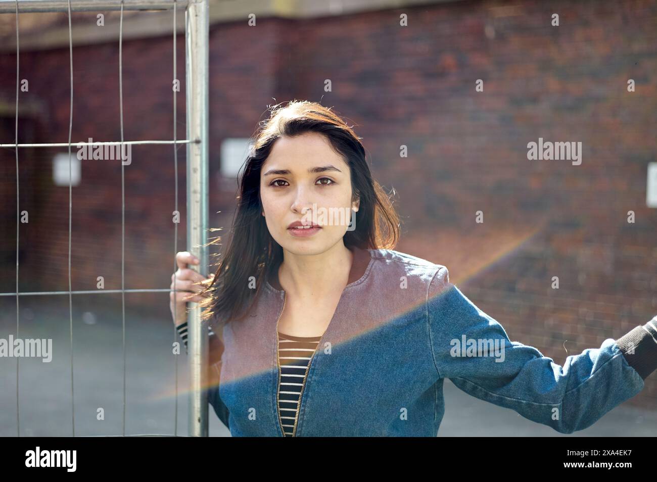A woman stands outdoors holding onto a chain-link fence, with a red ...