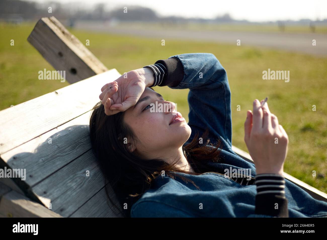 A woman is reclining on a wooden bench outdoors, shielding her eyes ...