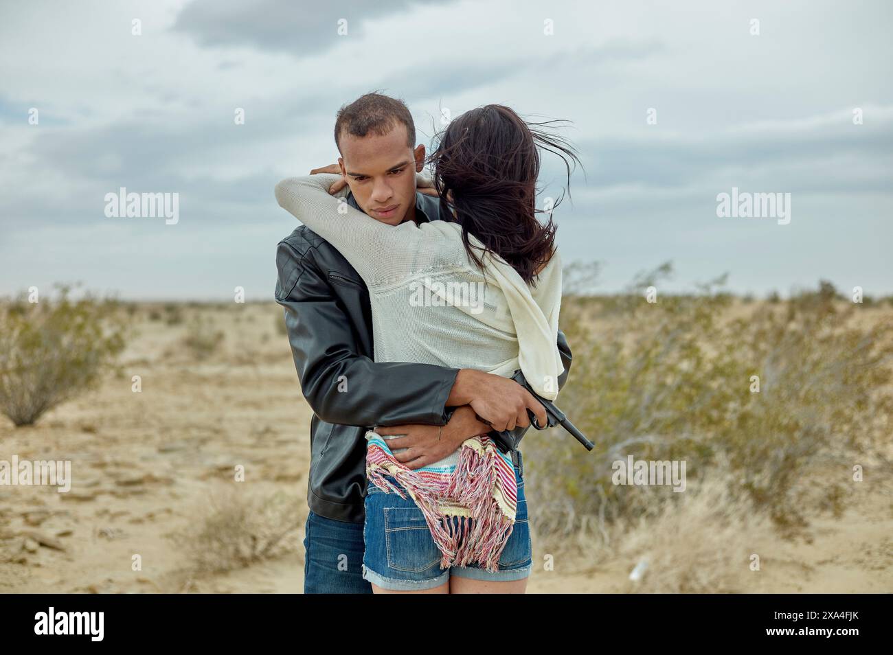 Two people embrace in a desert setting under an overcast sky, with ...