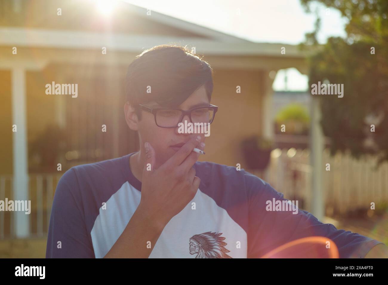 A young man stands outdoors smoking a cigarette during a sunset, with a ...