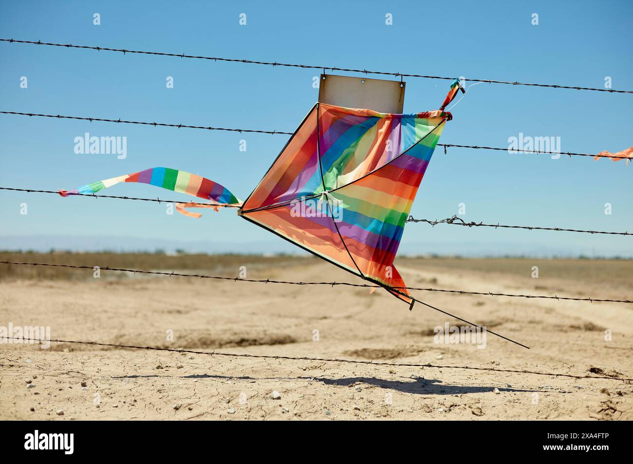 A colorful kite is entangled in a barbed wire fence against a clear ...