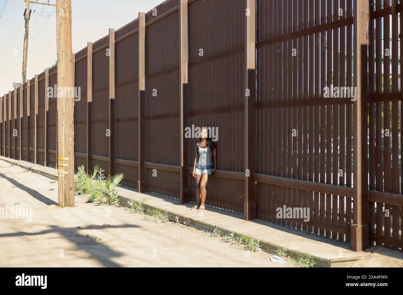 A woman stands by a tall metal fence on a sunny day, with a dirt path ...