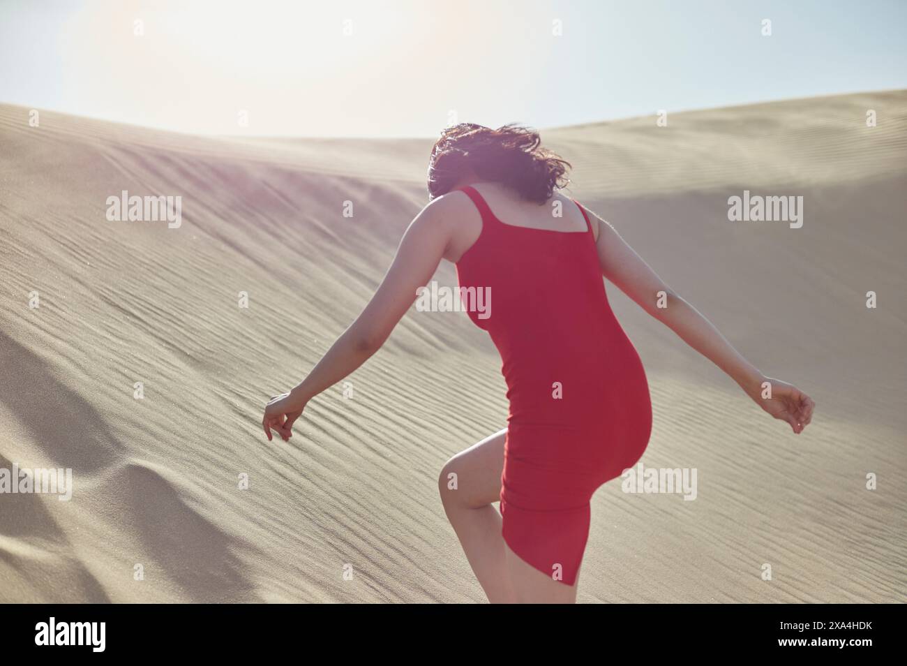 A woman in a red dress is walking up a sandy dune under bright sunlight ...