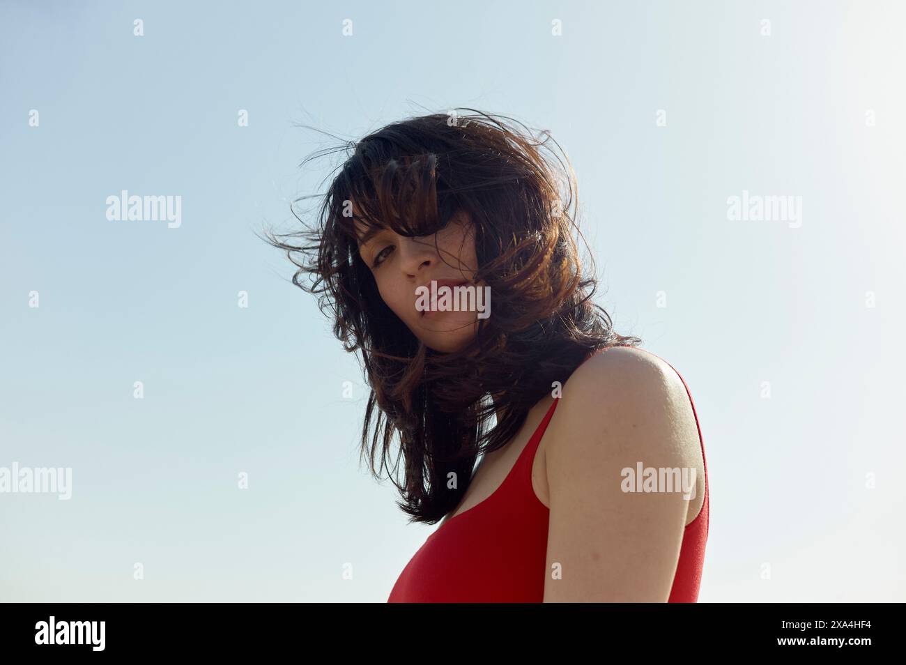 A woman wearing a red sleeveless top stands against a clear sky, with ...