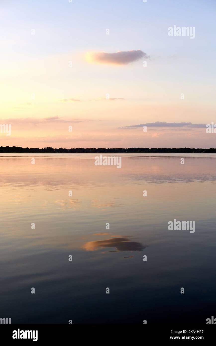 A serene lake scene at dusk with a single, small cloud reflected on the water's calm surface ...