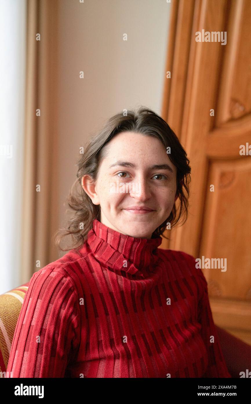A smiling woman in a red turtleneck sweater is seated indoors beside a ...