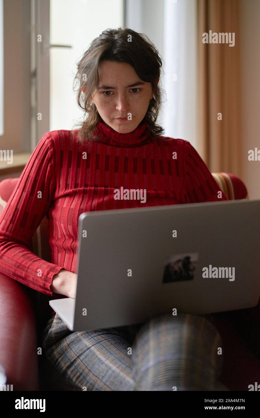 A woman is sitting on a chair indoors, intently looking at the screen ...