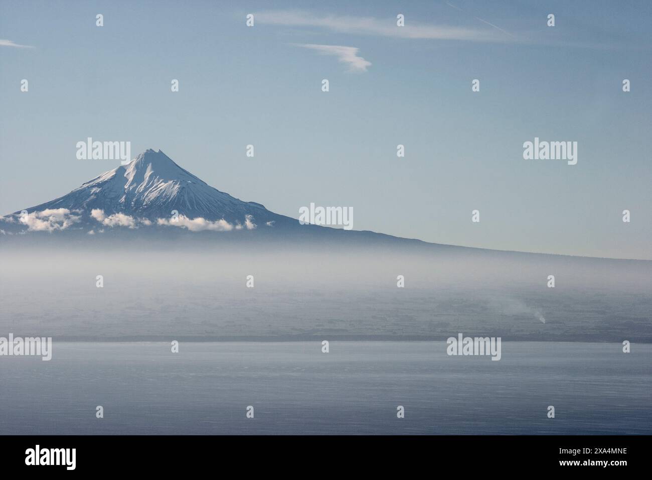 A majestic snow-capped mountain peak emerges above a blanket of clouds under a clear blue sky ...