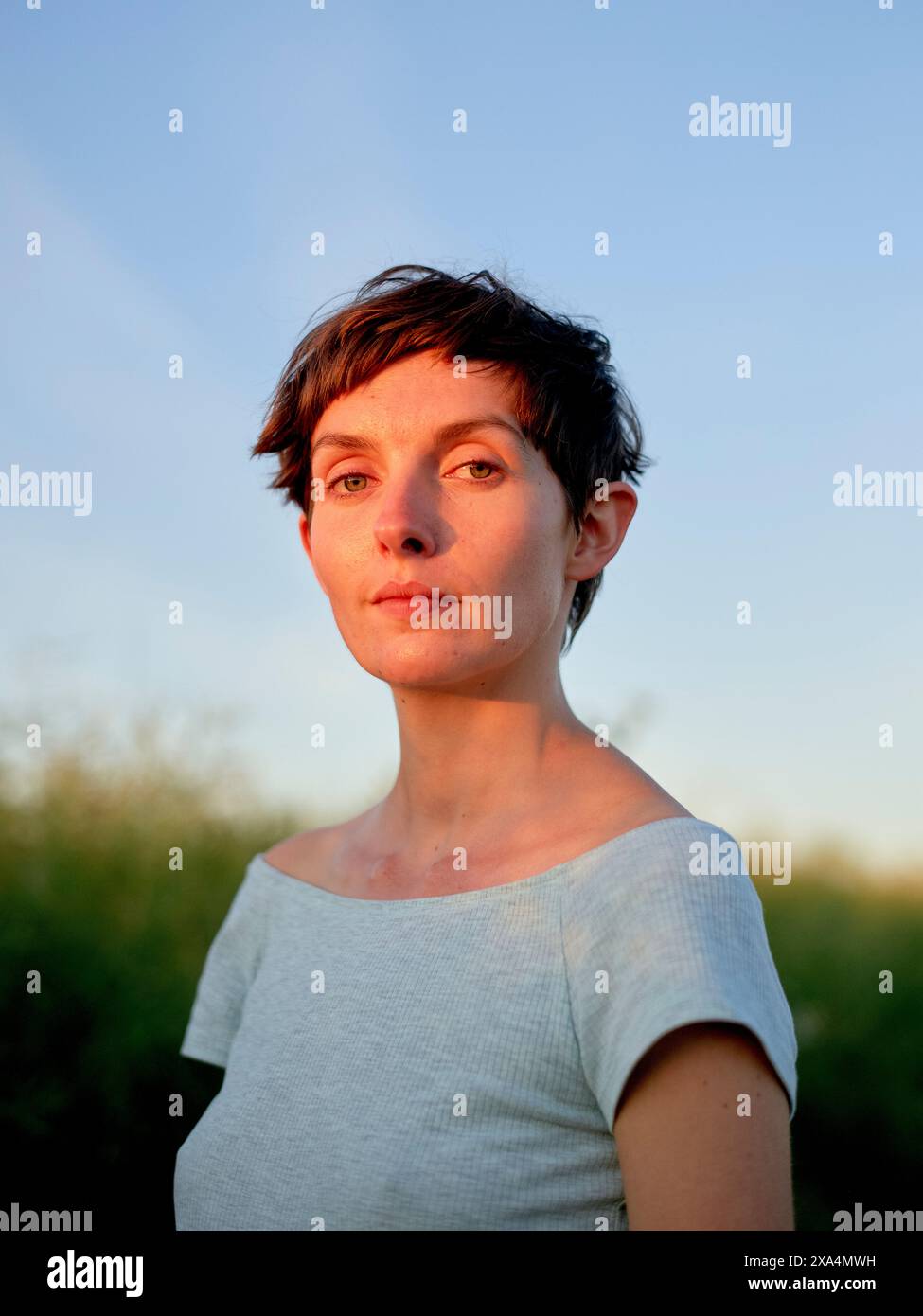 A woman with short hair stands outdoors during golden hour, her gaze ...