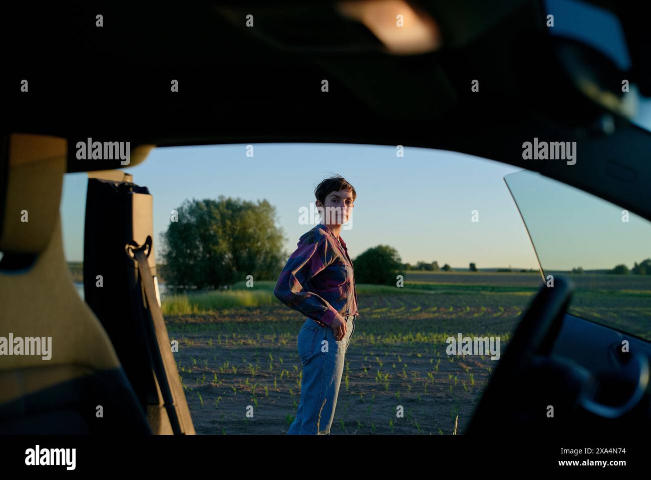 An young woman stands in a field viewed through the open rear window of a car during twilight ...