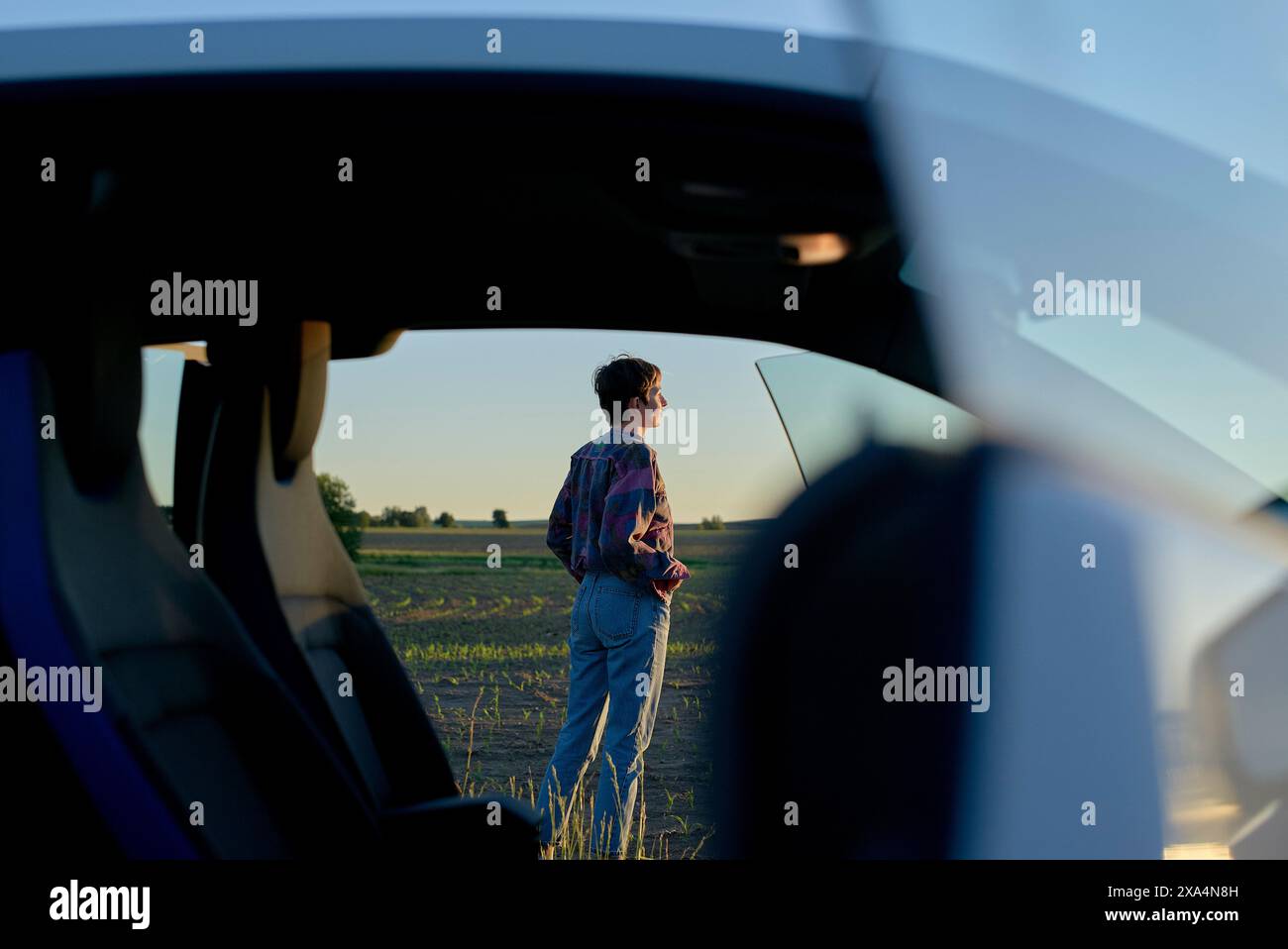 A young woman stands beside a parked car in a field at twilight, gazing ...