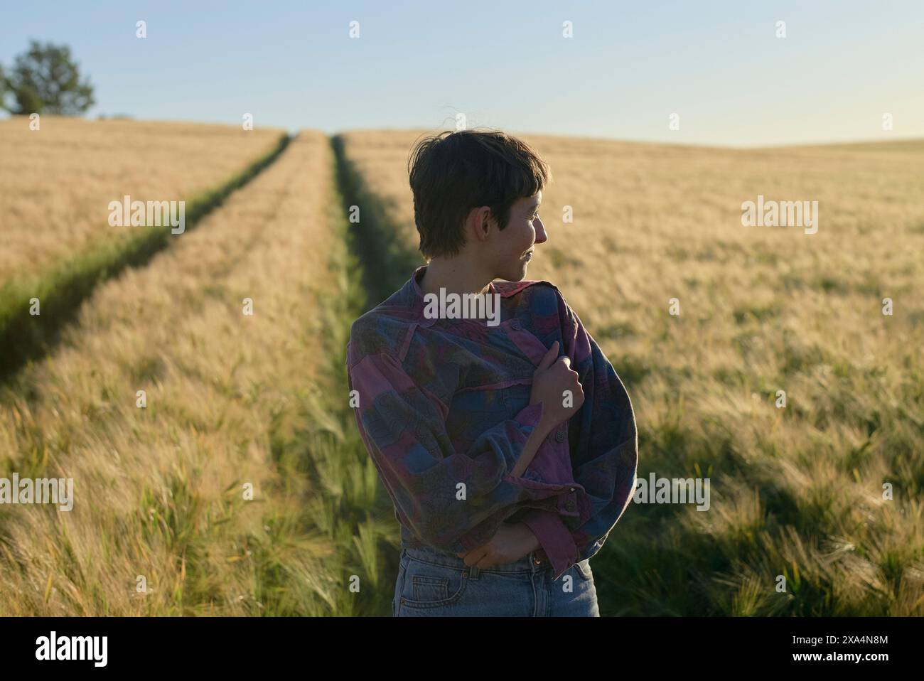 A young woman stands in a golden wheat field during sunset, looking ...