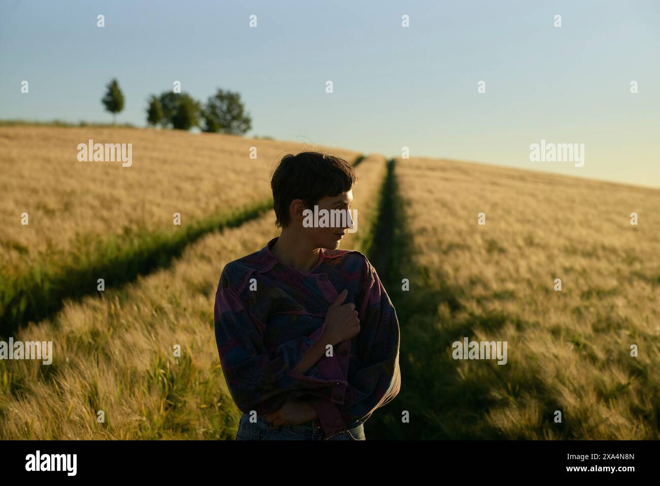 A young woman stands in a golden field at sunset, looking over their ...