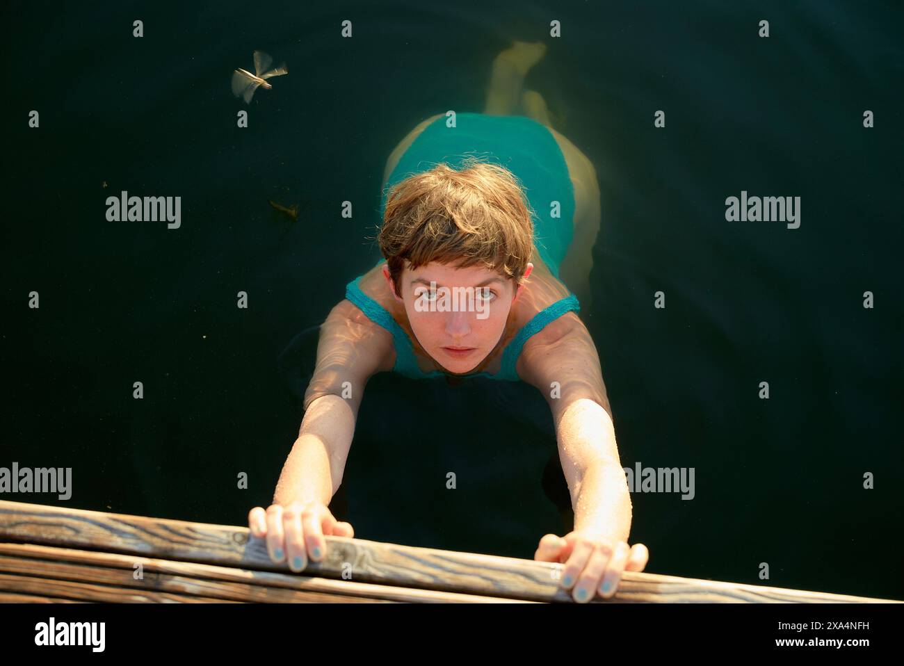 A young woman is gripping the edge of a wooden dock, peering up as they ...
