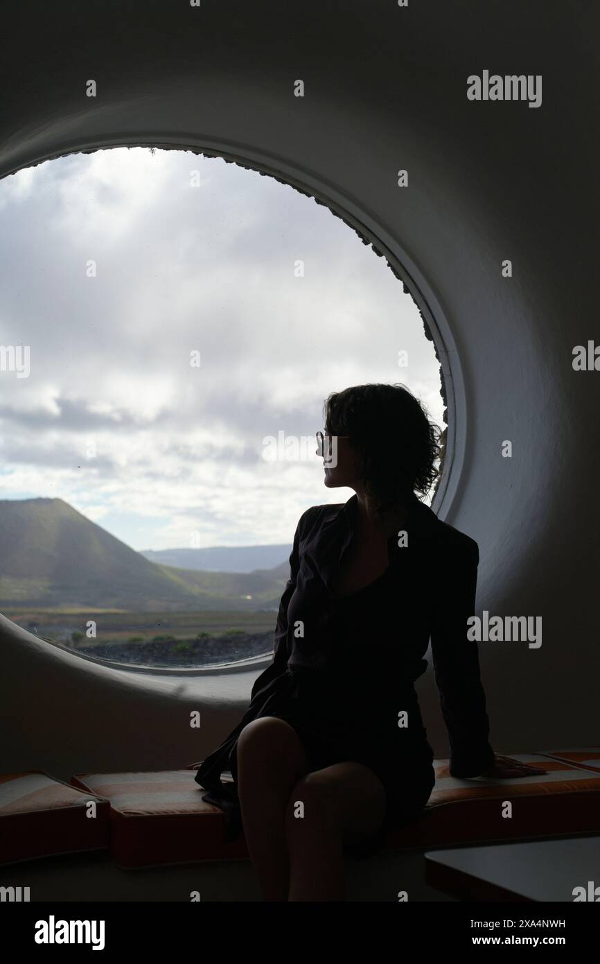 A silhouette of a young woman sitting by a round window with a view of ...