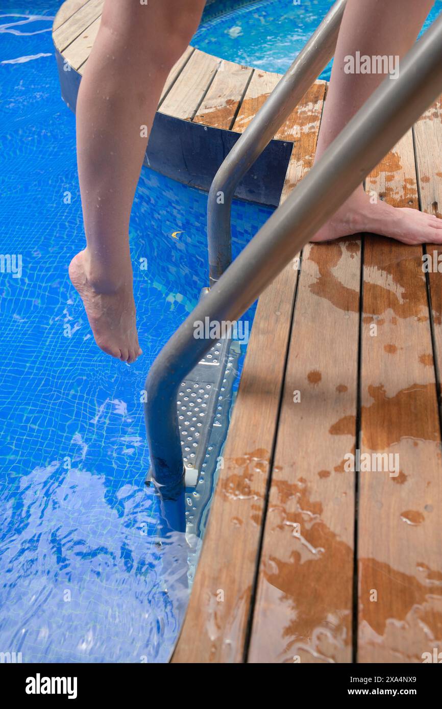 A young woman is dipping their toes into a swimming pool from a wooden ...