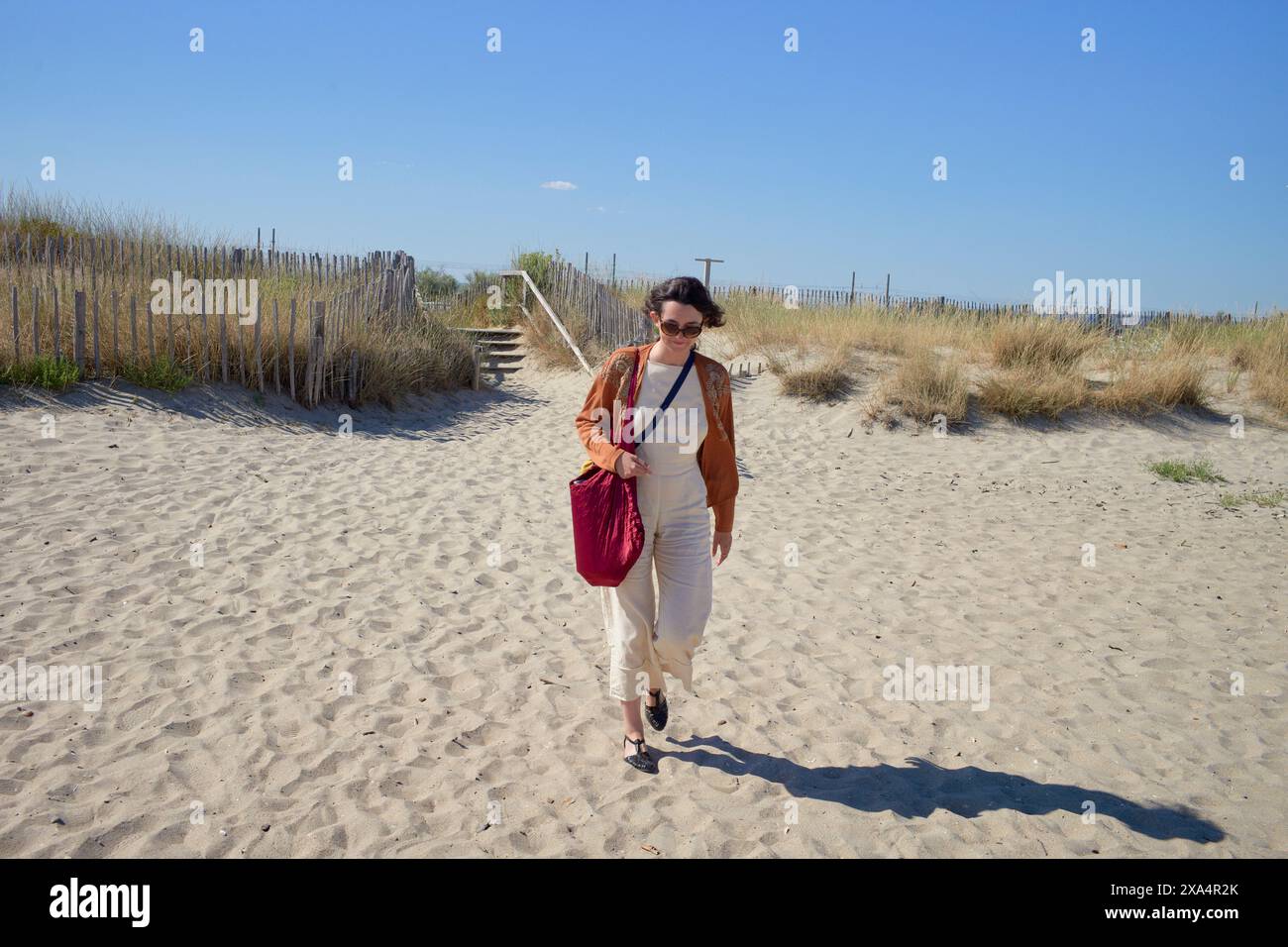 A woman is walking on a sandy beach path with dunes and a wooden fence ...