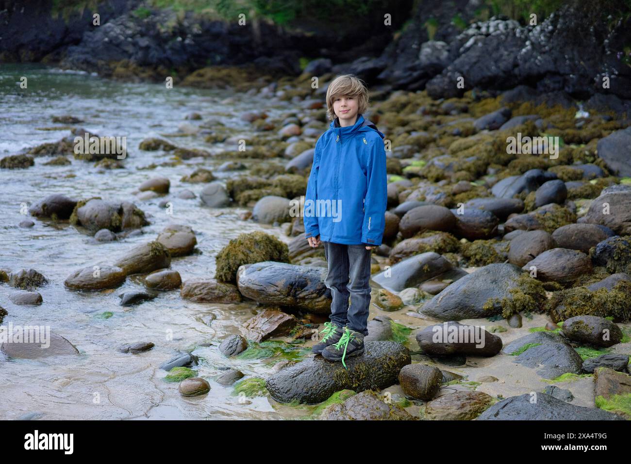 Young boy standing on a rocky shoreline with seaweed and moss-covered ...