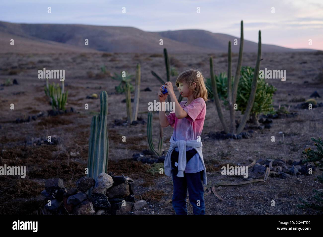 A Young Girl With Blonde Hair Takes A Photo With A Compact Camera In A Desert Landscape At Dusk