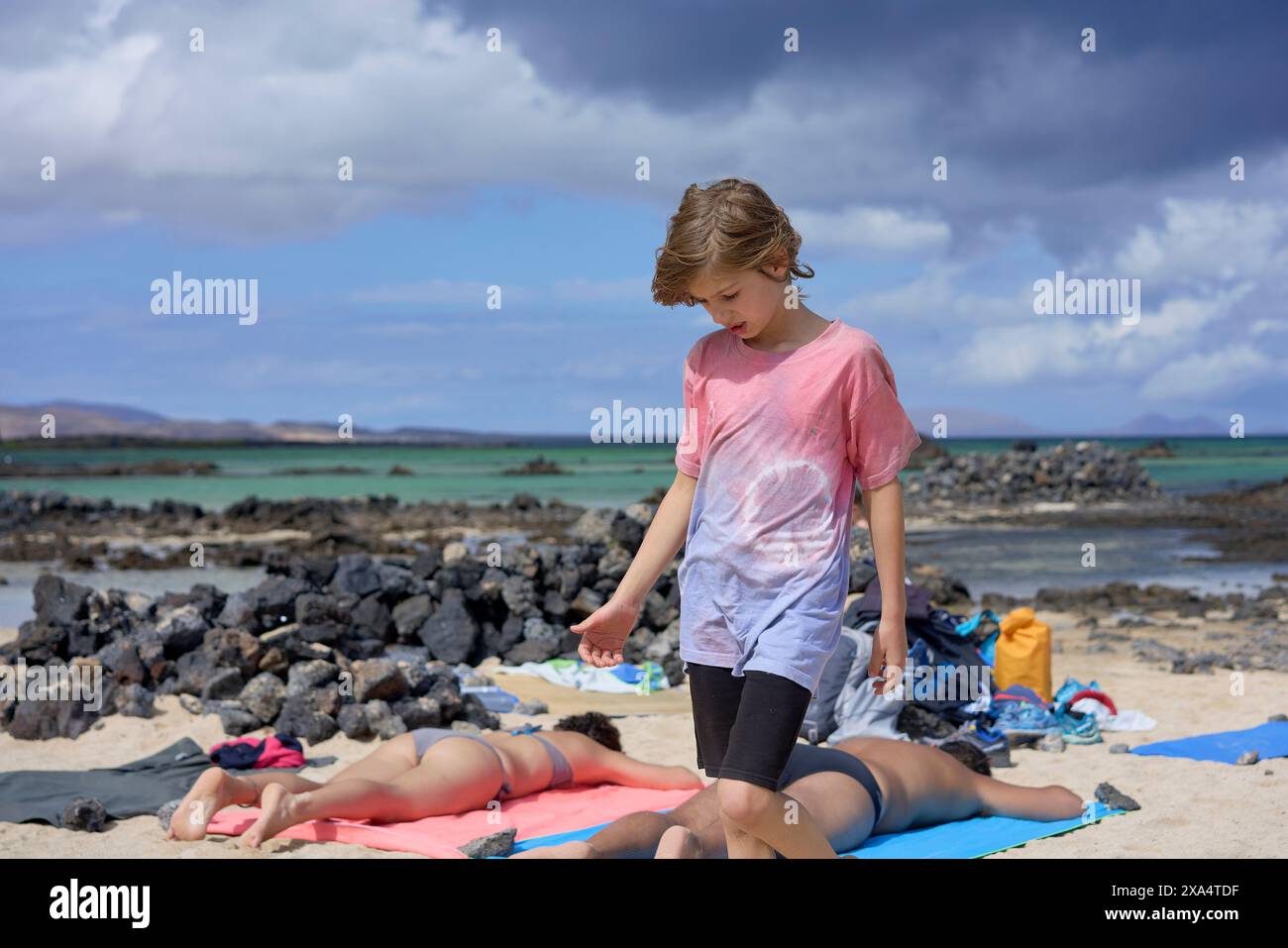 Young boy walking by two people sunbathing on a beach with dark clouds ...
