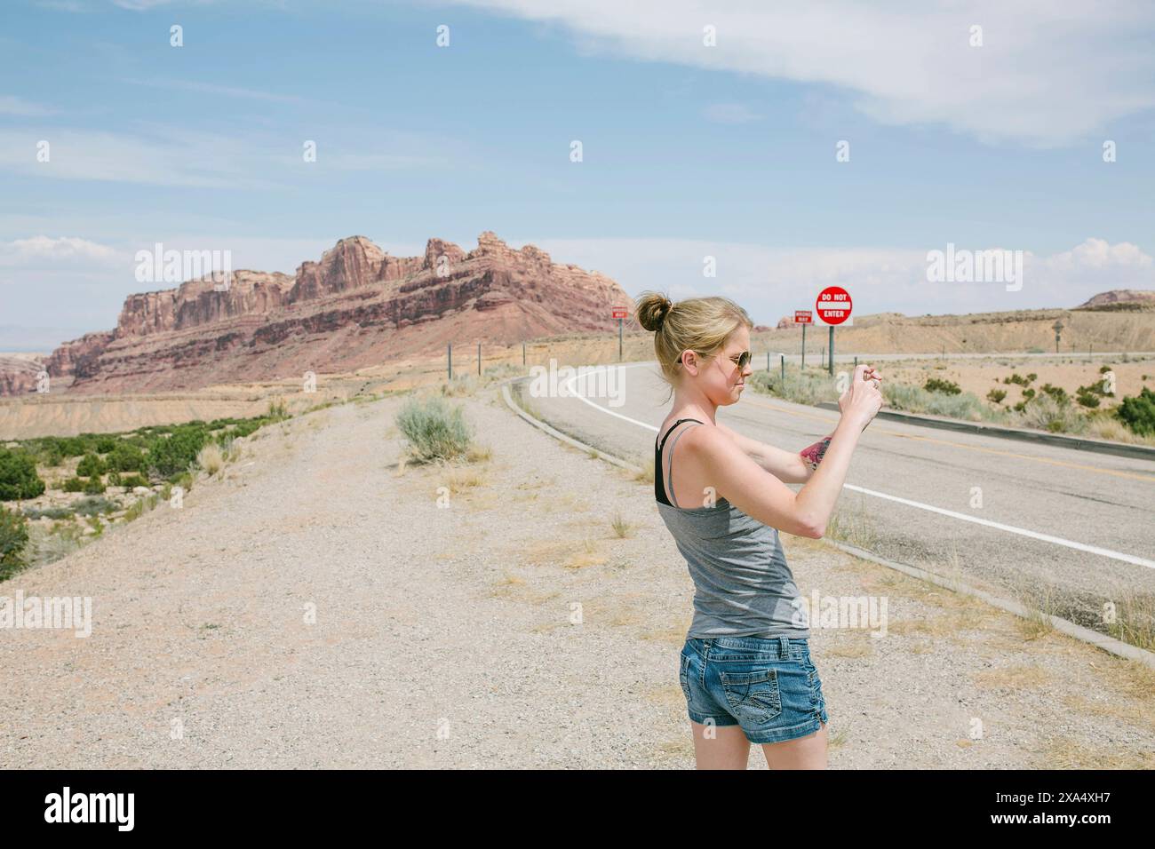Woman hitchhiking on a deserted road with a clear blue sky and rocky mountains in the background ...