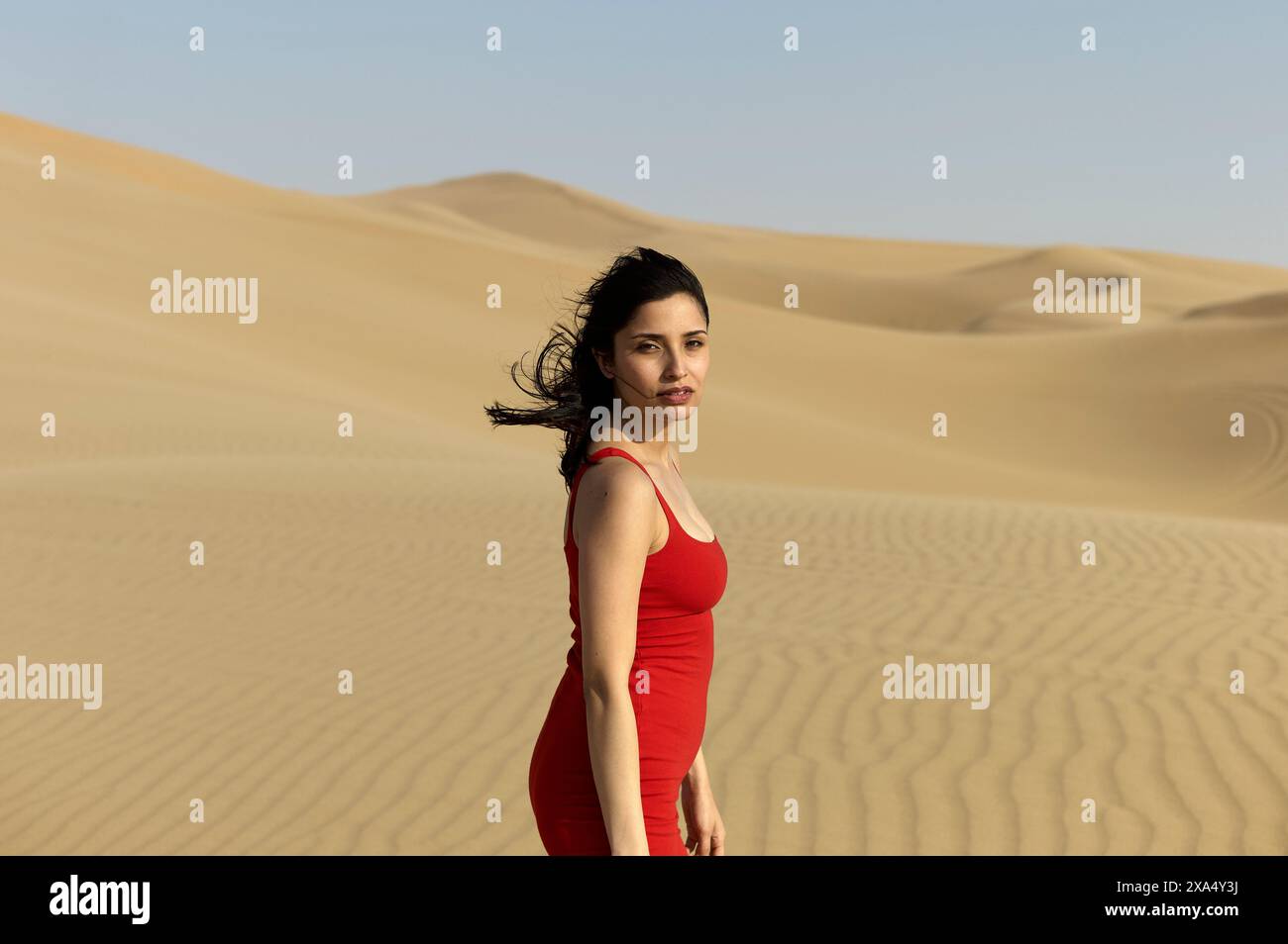 Woman in a red dress standing in a desert with sand dunes under a clear ...