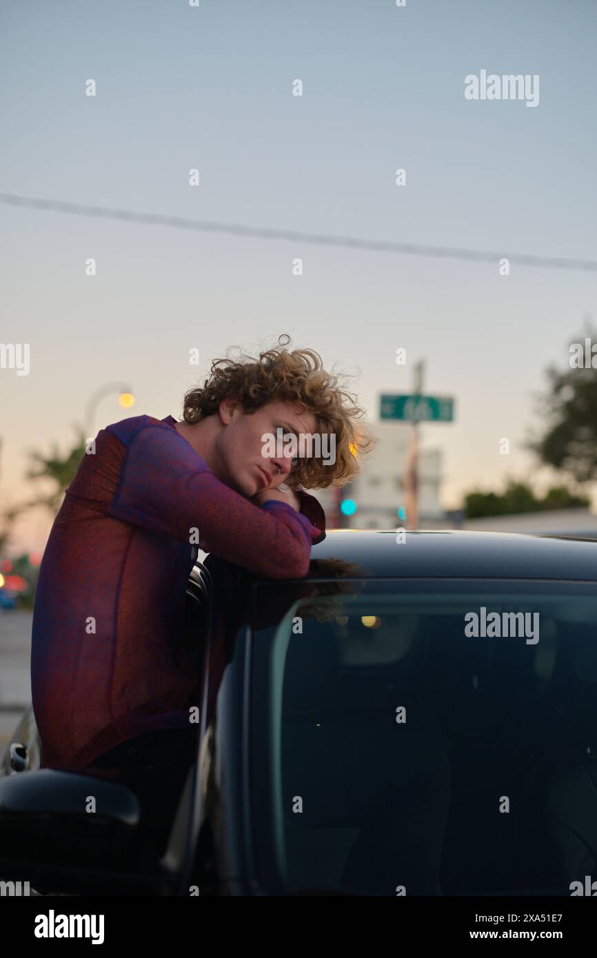 Young curly-haired man leaning on a car at dusk with city lights in the ...