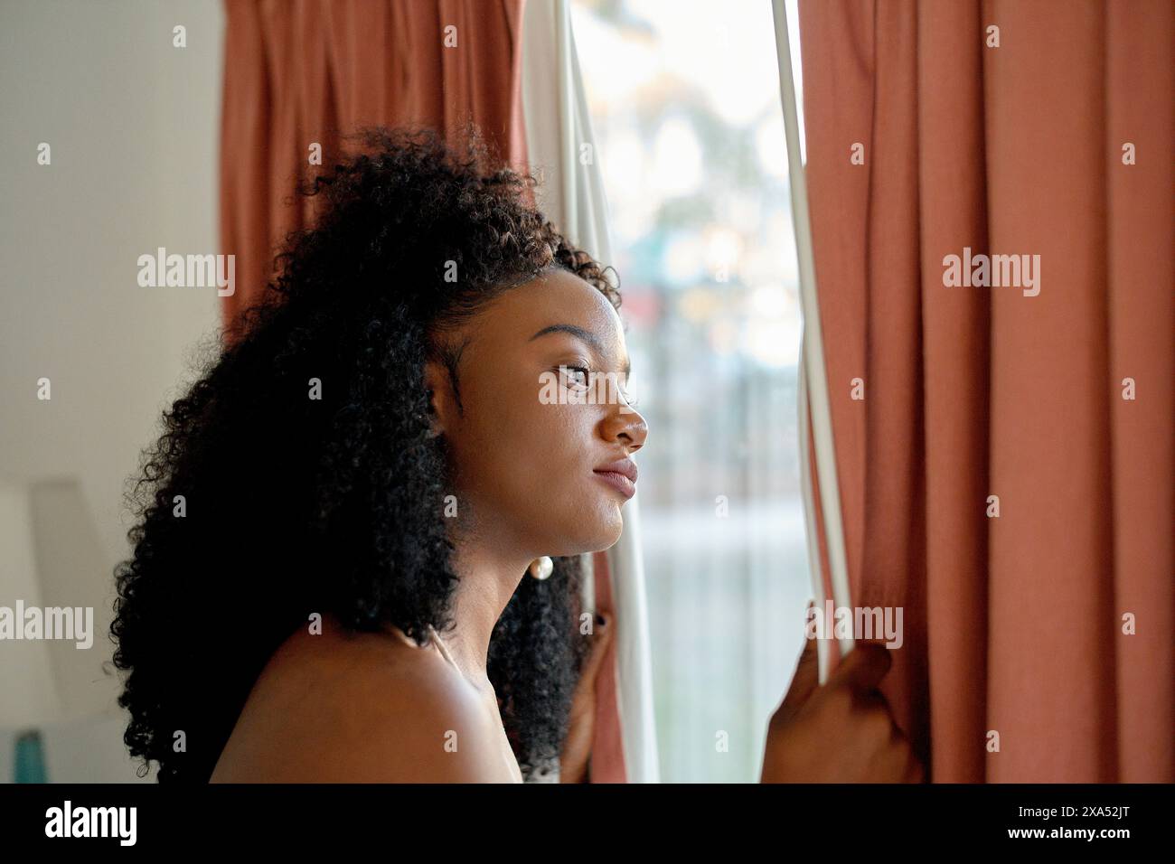 Woman gazing out a window with curtains, displaying a thoughtful ...