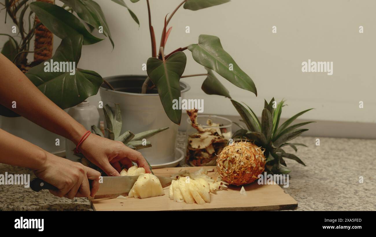 A person is slicing a pineapple on a cutting board next to a potted ...